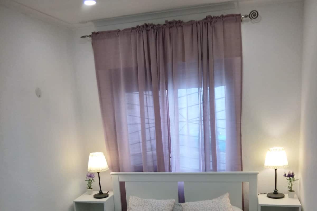 Uma cama bem arrumada com almofadas e um cobertor, duas mesas de cabeceira com abajures, uma janela com cortinas e uma cabeceira.