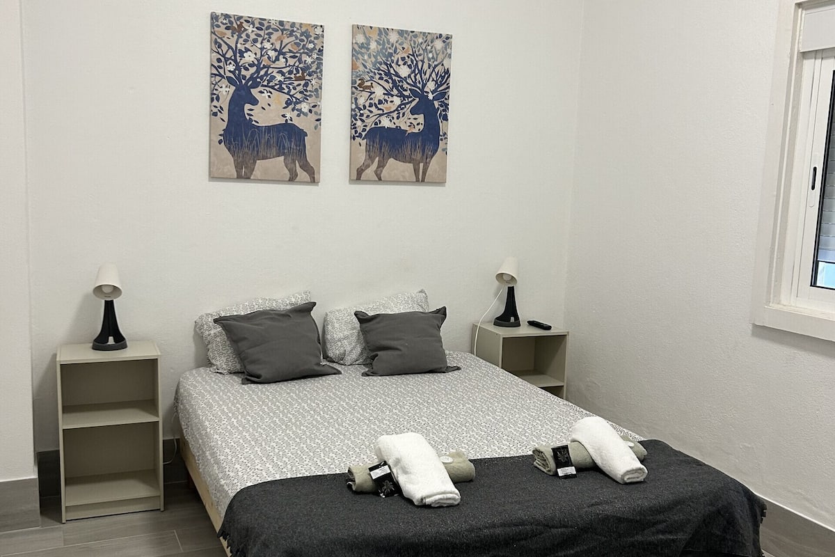 Quarto bem arrumado com cama, duas mesas de cabeceira, uma janela e duas obras de arte na parede.