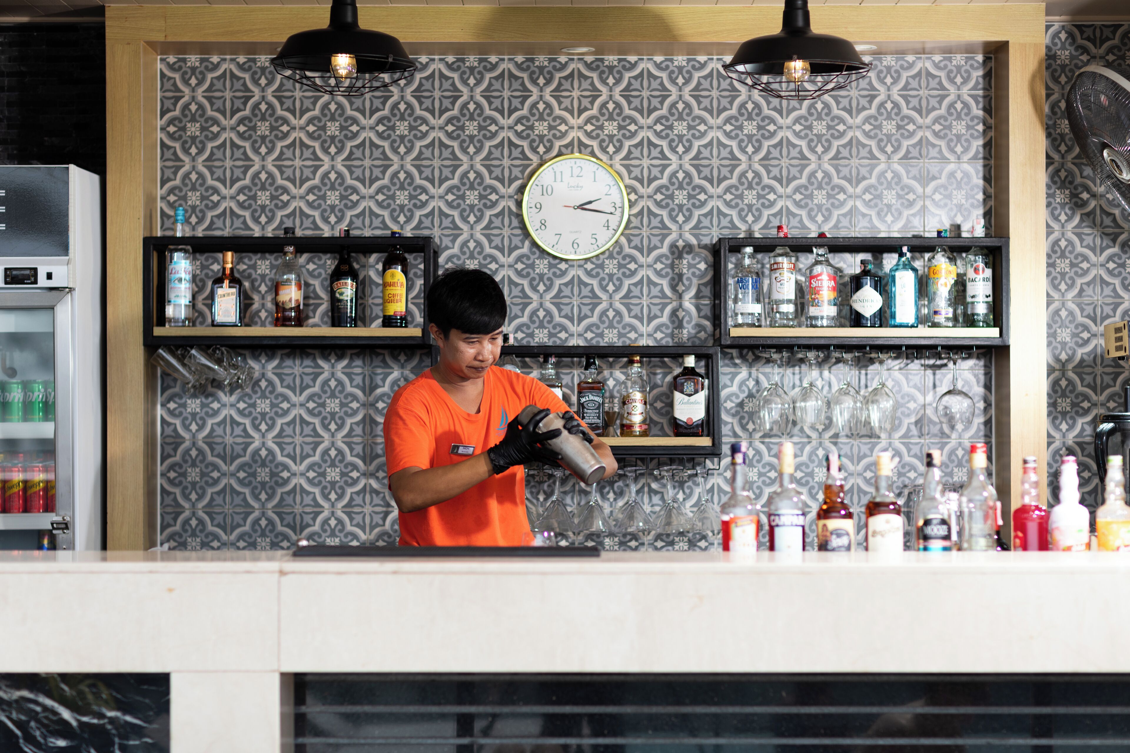 Uma área de bar com prateleiras de garrafas de bebidas, um relógio e uma pessoa preparando uma bebida.