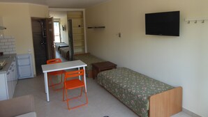 Quarto compacto com cama, escrivaninha com cadeira, televisão e área de chuveiro.