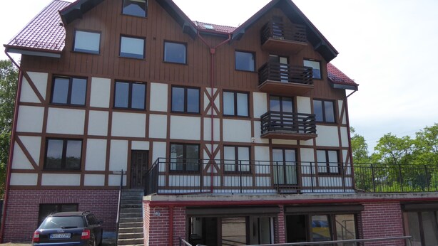 Um edifício de dois andares com estilo arquitetônico tradicional, combinando construção em madeira e tijolo, com uma varanda no andar superior e um carro estacionado na frente.
