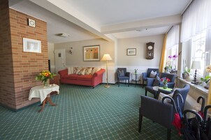 Quarto de hotel com parede de tijolos, um sofá vermelho, uma poltrona azul, uma pequena mesa com abajur e uma janela com plantas.