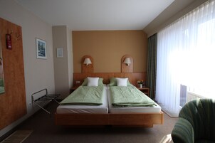 Quarto de hotel com duas camas, cabeceira de madeira, mesa de cabeceira com abajur, uma cadeira e uma janela com cortinas.