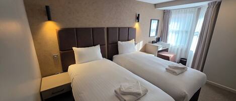 Quarto de hotel com duas camas, uma mesinha de cabeceira, escrivaninha e janela com cortinas.