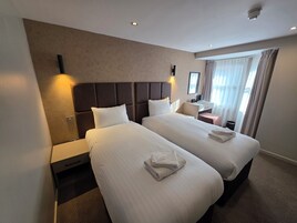 Quarto de hotel com duas camas, uma mesinha de cabeceira, escrivaninha e janela com cortinas.