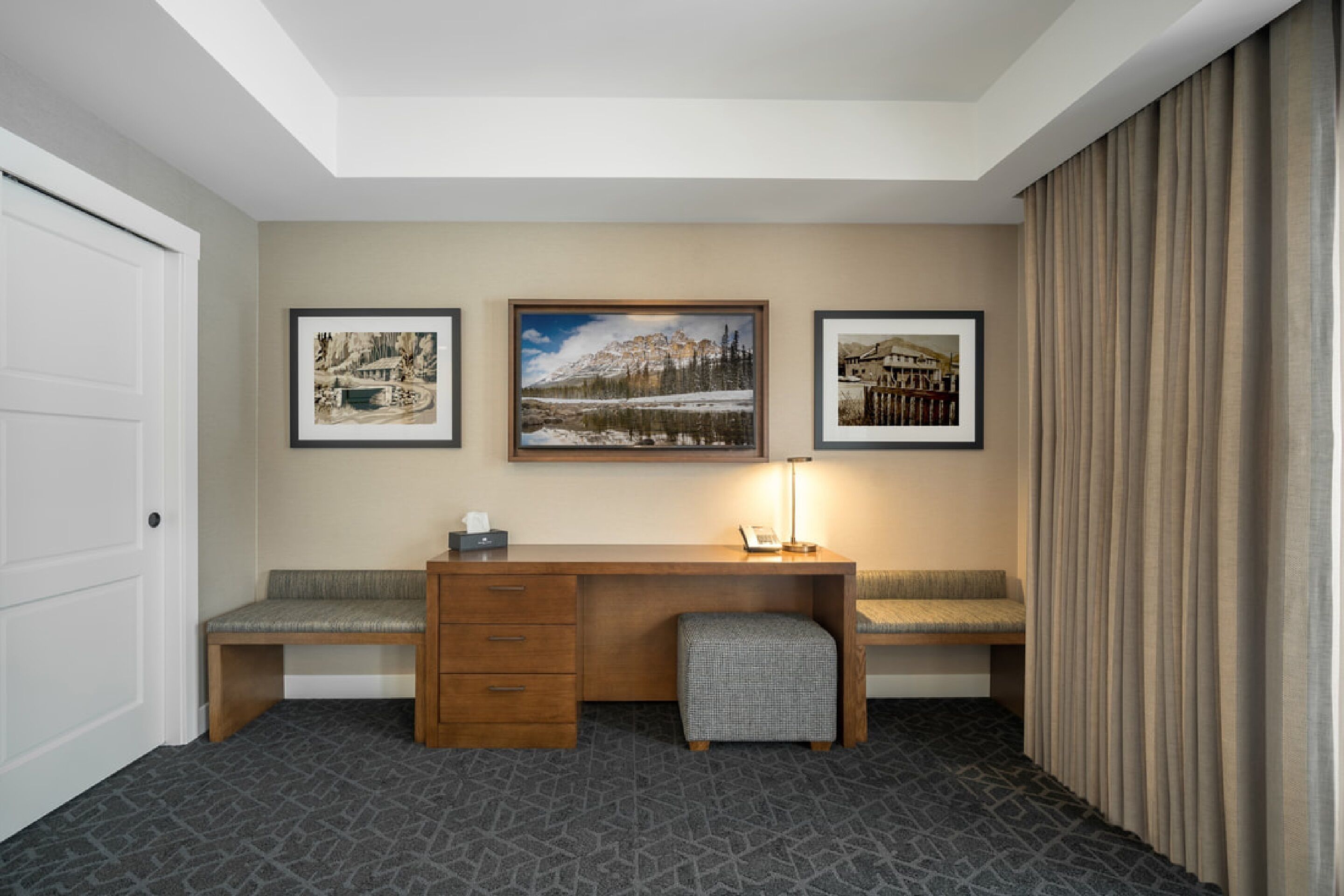 Quarto de hotel com escrivaninha de madeira, dois bancos acolchoados, um pequeno banquinho e três quadros na parede.