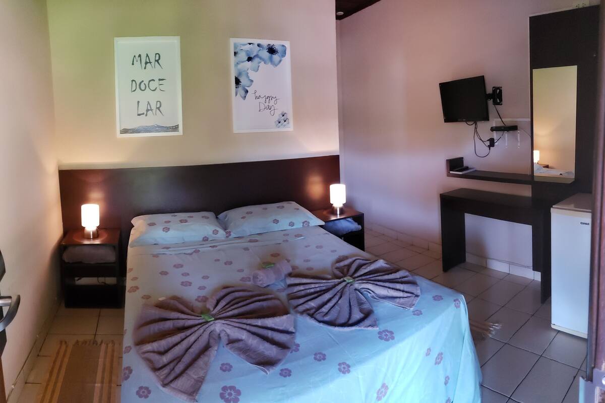 Quarto de hotel com cama, duas mesas de cabeceira com abajures, televisão de tela plana na parede e um refrigerador.