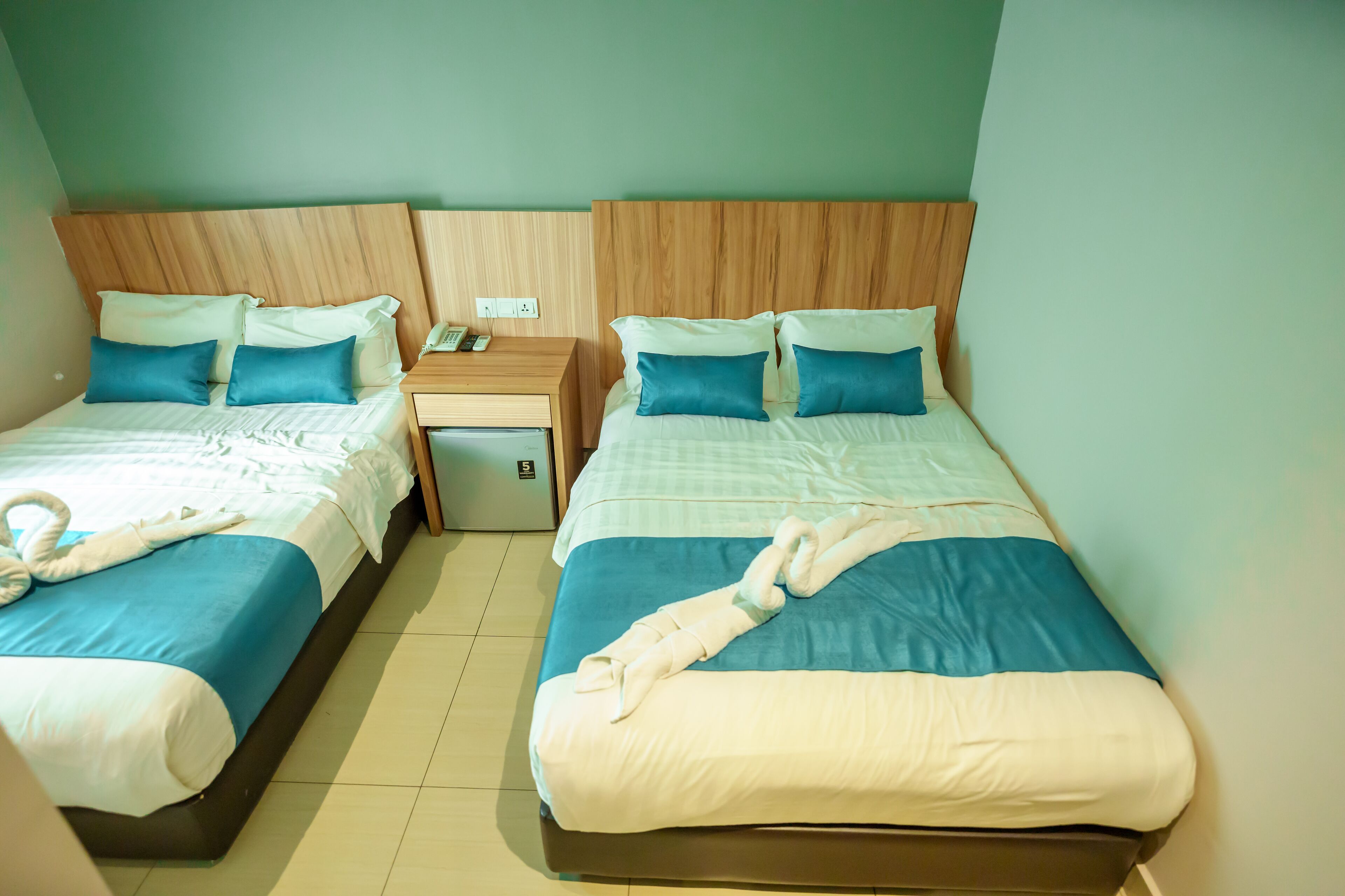 Quarto de hotel com duas camas, cada uma com roupa de cama azul e branca, e cabeceira de madeira. Há uma pequena mesa de cabeceira com telefone e tomada.
