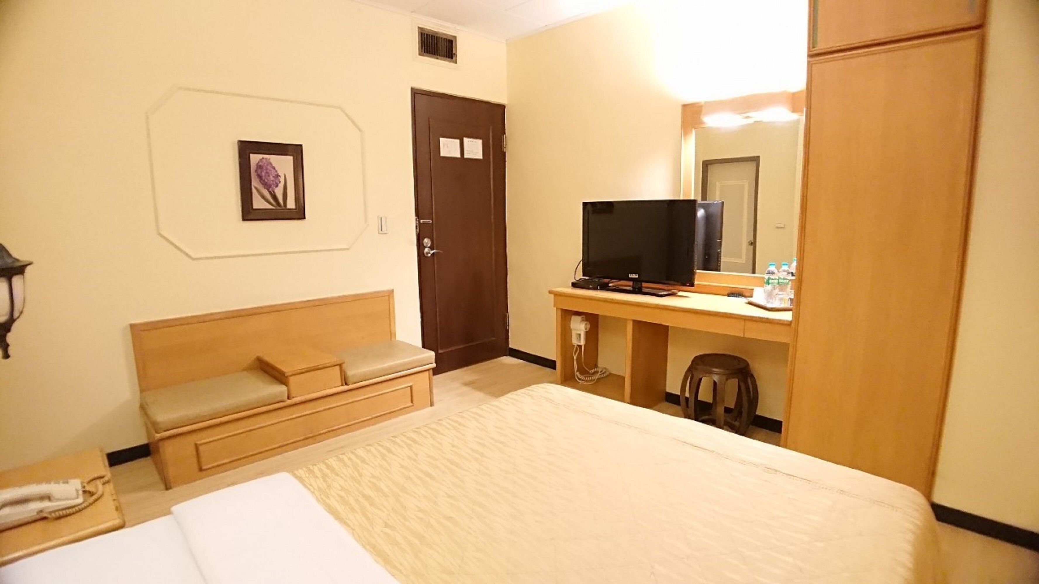 Quarto de hotel com cama, escrivaninha com televisão, um banco de madeira, um banquinho e uma luminária fixada na parede.