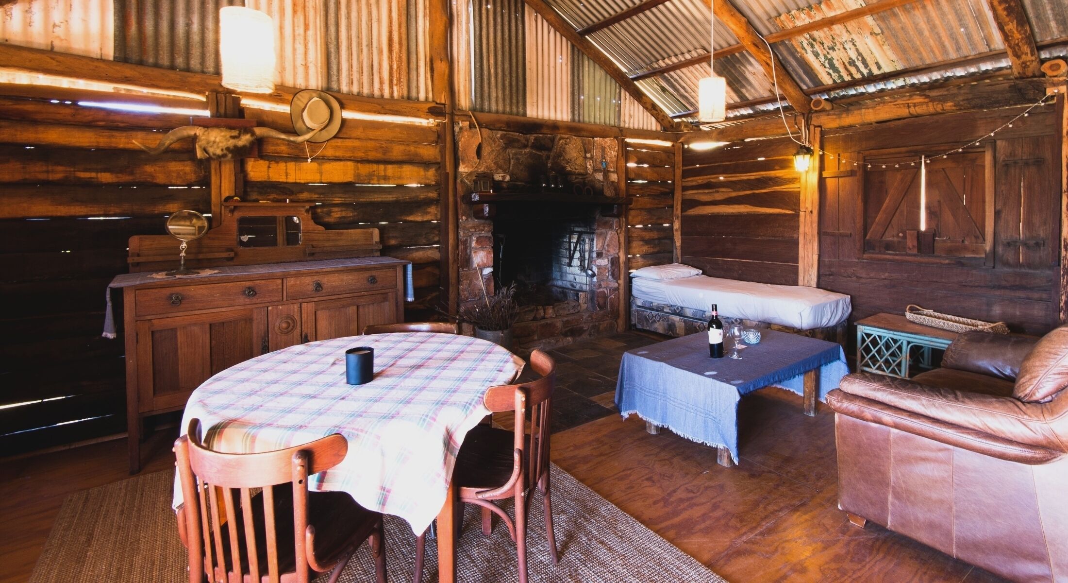 Interior rústico de cabana com mesa de jantar, cadeiras, sofá, pequena mesa com garrafa e copo, e lareira de pedra.