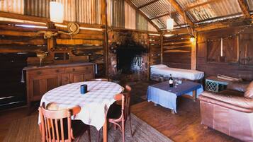 Interior rústico de cabana com mesa de jantar, cadeiras, sofá, pequena mesa com garrafa e copo, e lareira de pedra.