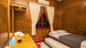 Um quarto pequeno e acolhedor com paredes de madeira, uma cama de solteiro, uma escrivaninha com telefone e caixa de lenços, e uma janela com cortinas.