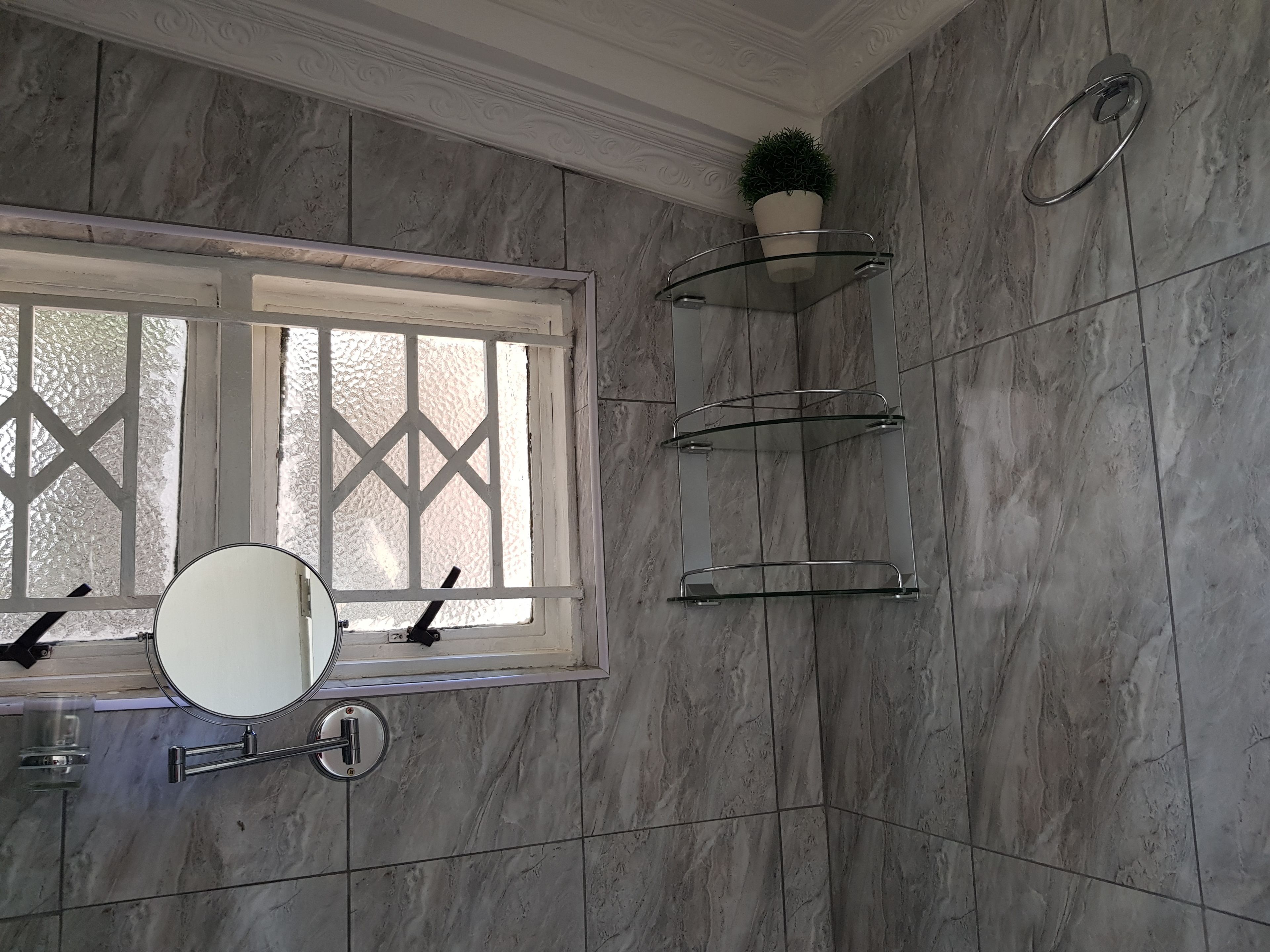 Um banheiro com paredes de mármore, uma janela com grade decorativa, um espelho redondo e uma prateleira de vidro com planta em vaso.