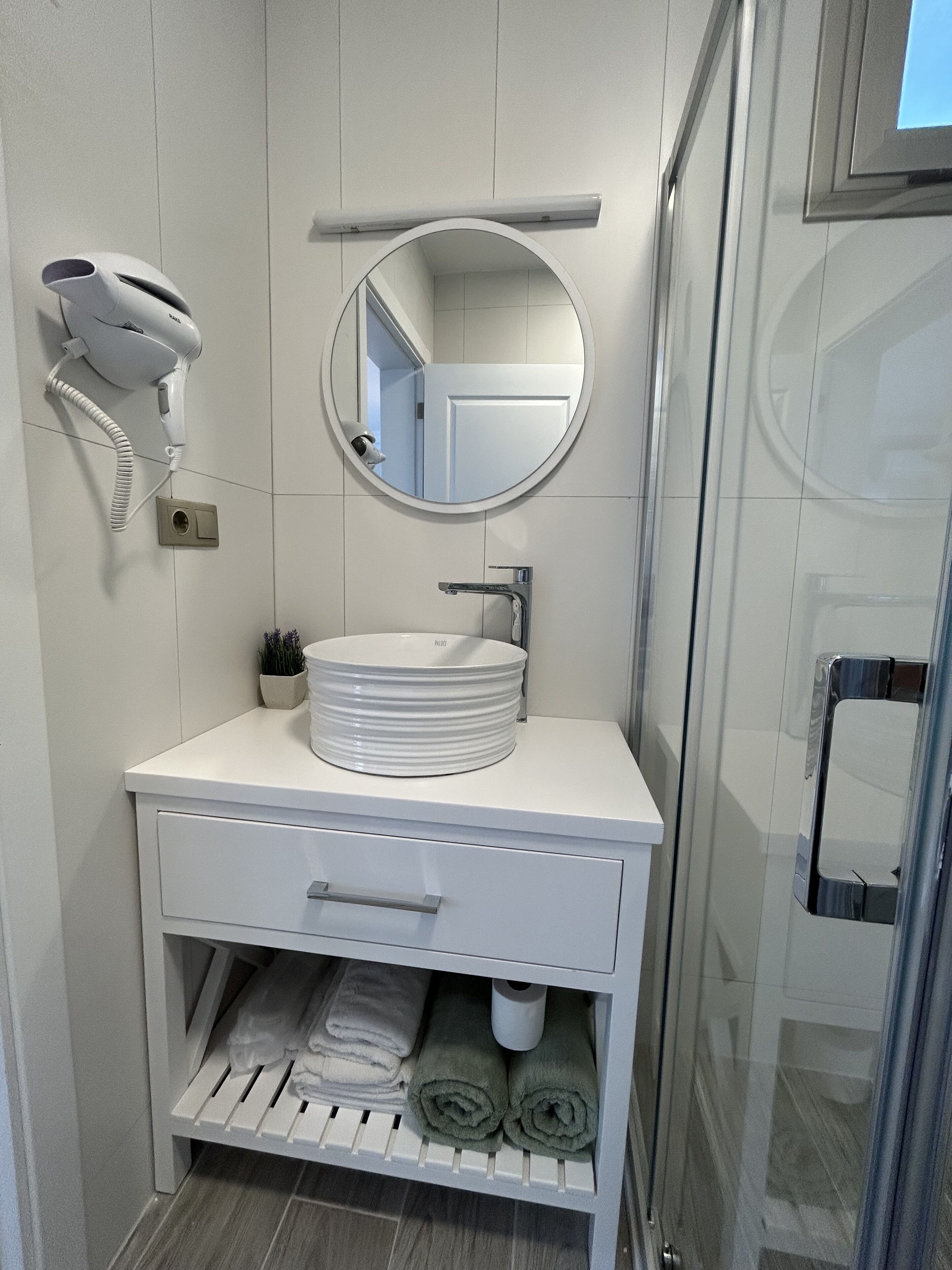 Uma casa de banho moderna com um espelho redondo, uma pia e um secador de cabelo fixado na parede.