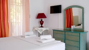 Cama bem arrumada com lençóis brancos, abajur vermelho sobre uma mesa pequena, cômoda verde e um espelho grande.