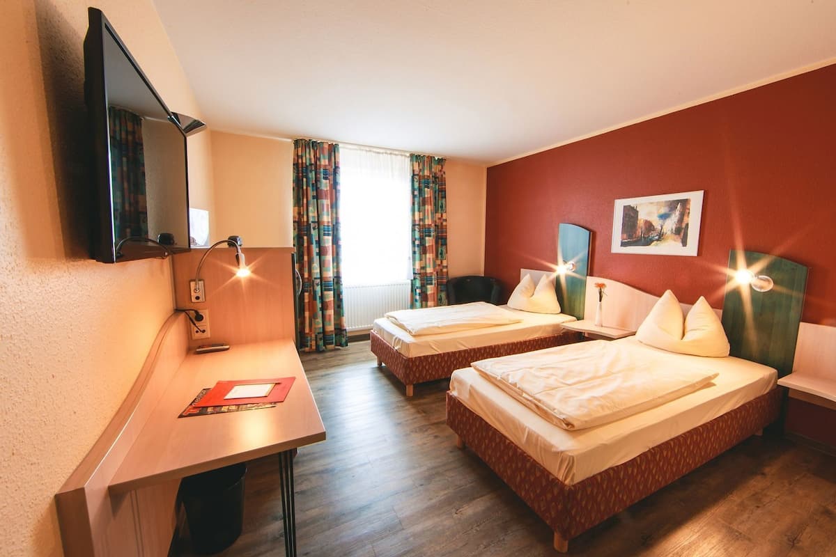 Quarto de hotel com duas camas, uma escrivaninha, televisão e janela com cortinas.