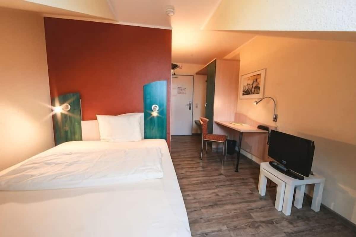 Quarto de hotel com cama, escrivaninha, cadeira e televisão sobre uma mesinha.