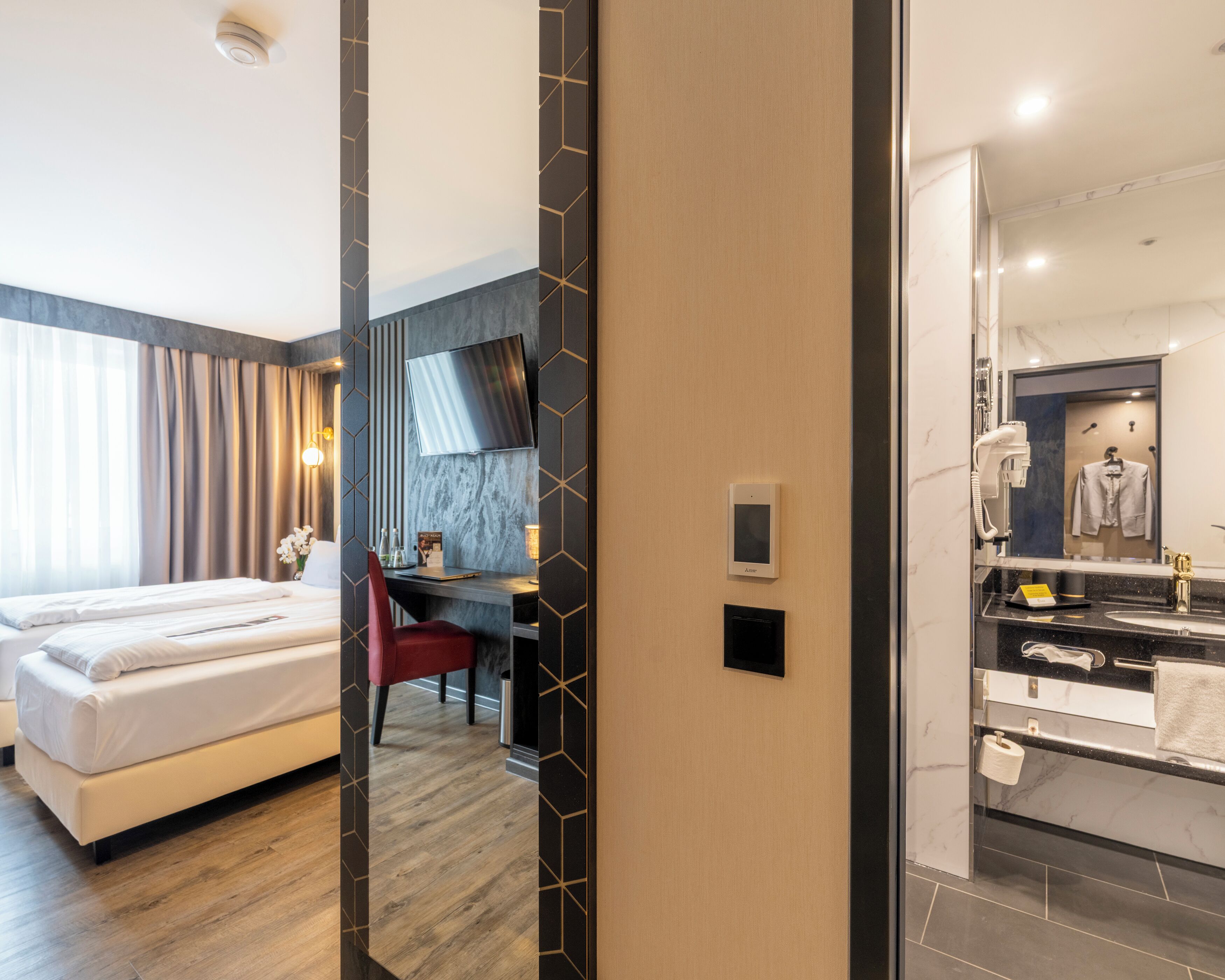 Quarto de hotel com cama, escrivaninha, cadeira, televisão e banheiro com espelho e pia.