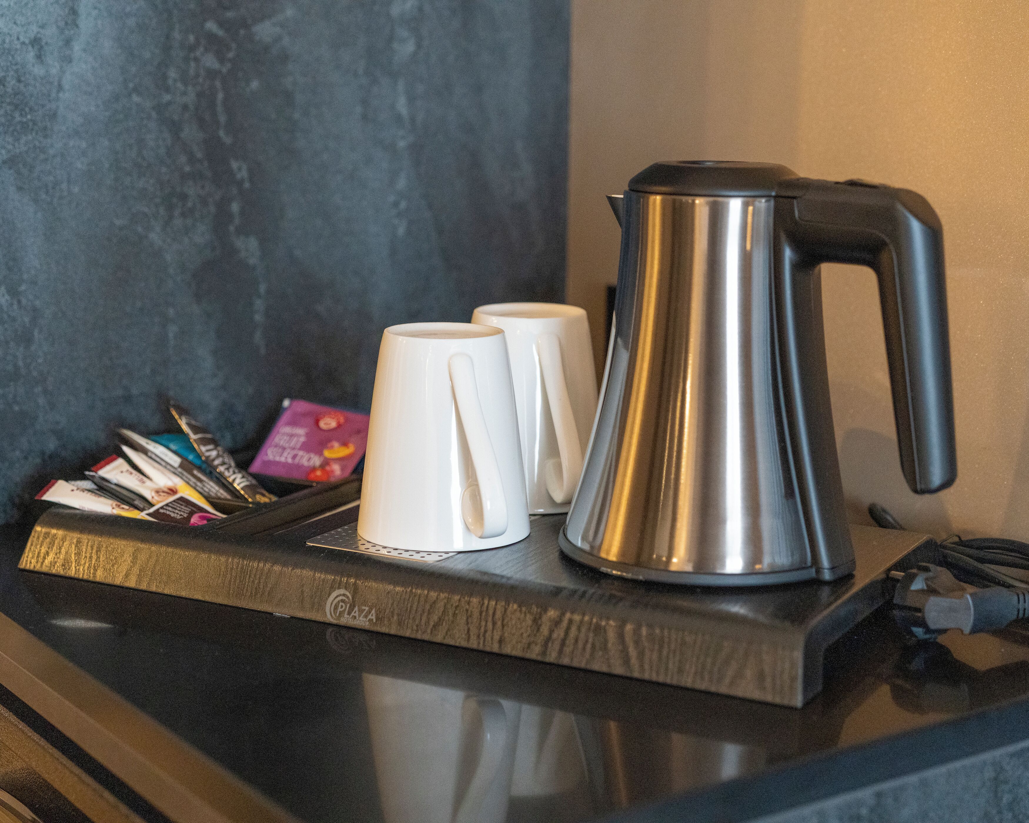 Um canto de café com um bule de aço inoxidável, duas canecas brancas e uma bandeja com revistas e uma caixa rosa.