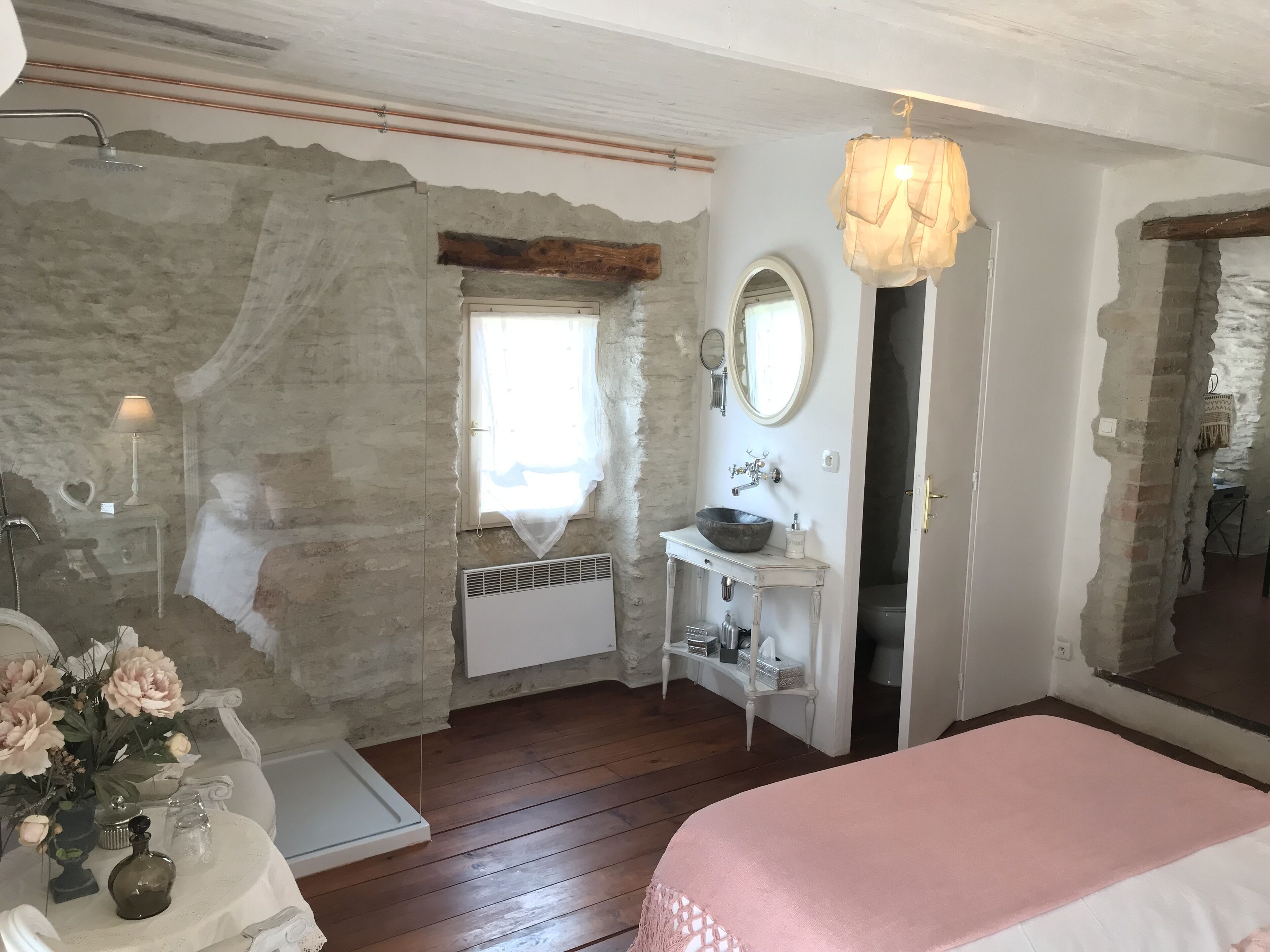 Um quarto com parede de pedra, piso de madeira, uma cama com manta rosa, mesa de cabeceira com abajur, cômoda, espelho e janela com vista para o exterior.