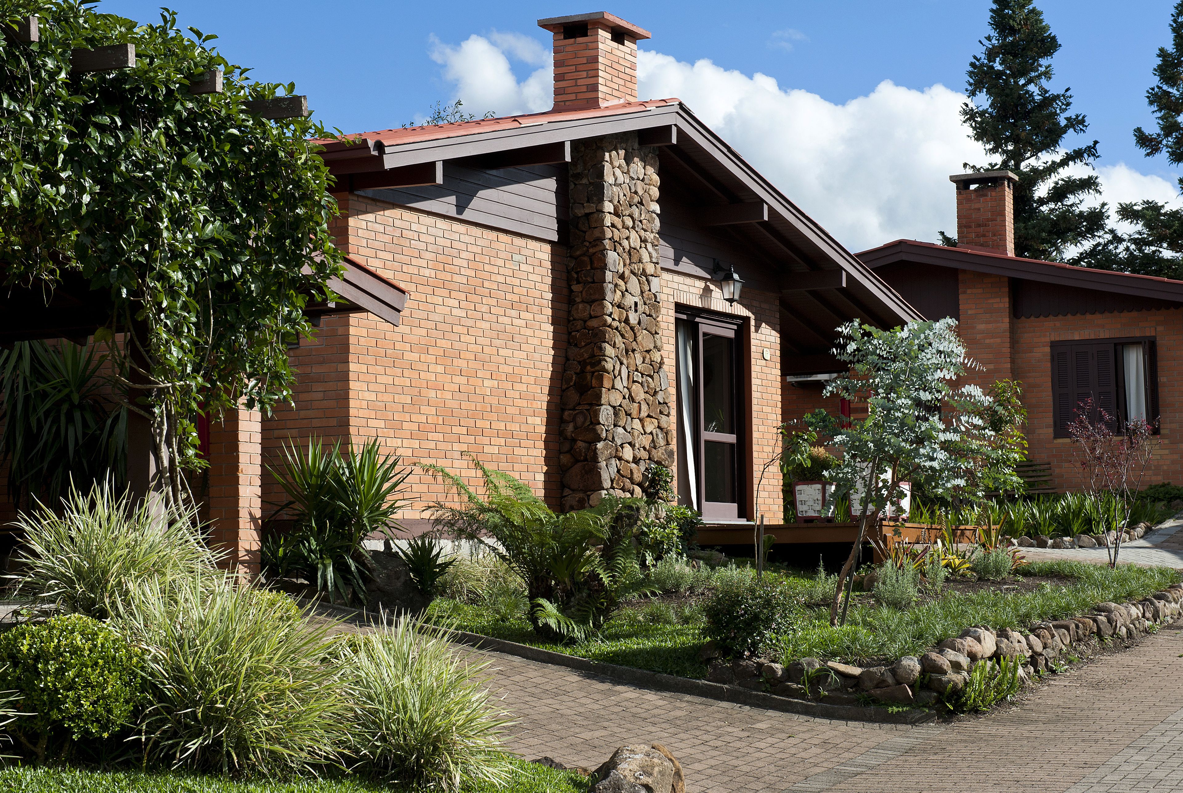 Uma casa de tijolos com chaminé de pedra, rodeada por um jardim com plantas variadas e um caminho pavimentado.
