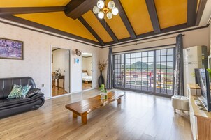 Uma sala de estar ampla com piso de madeira, um sofá de couro preto, uma mesa de centro de madeira e uma grande janela com vista para uma paisagem montanhosa.