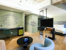 Quarto de hotel moderno com cama, televisão de tela plana, escrivaninha com cadeira e mesa de centro redonda.