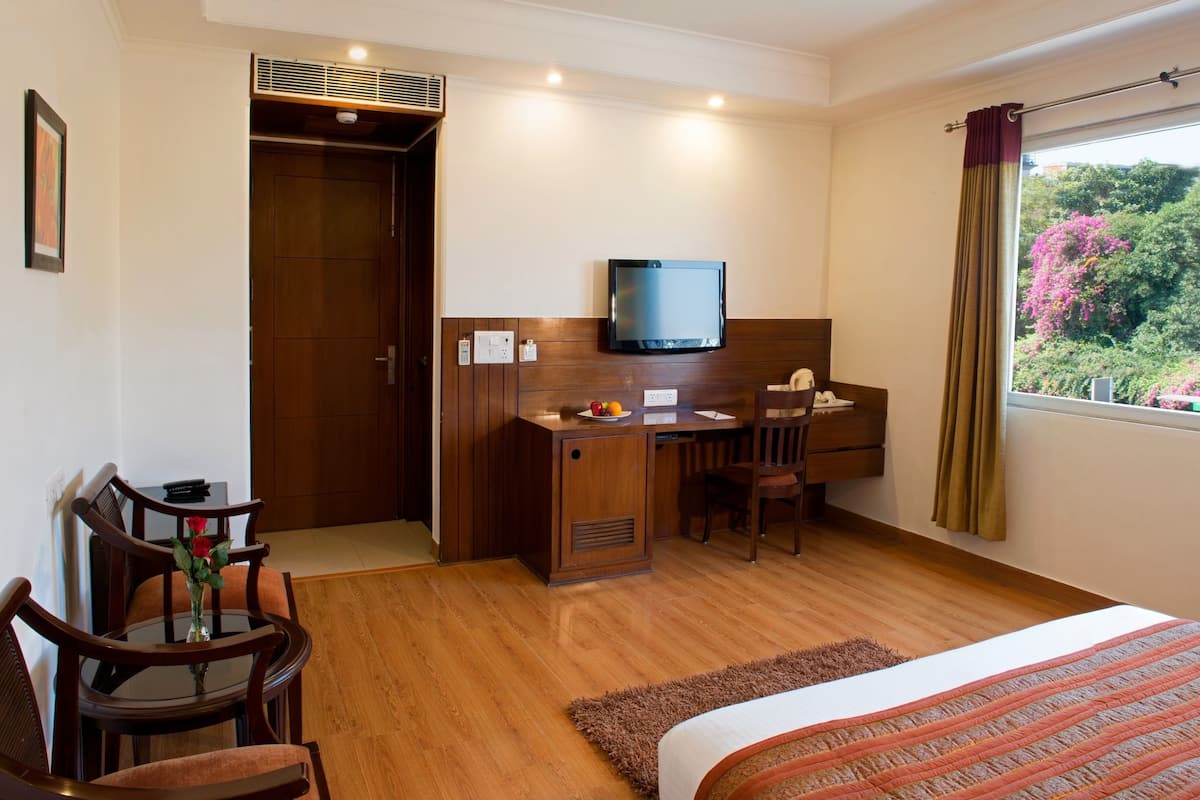 Quarto de hotel com escrivaninha de madeira, televisão, cama com colcha estampada e janela com cortinas.