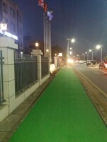 Uma passarela pavimentada com superfície verde, limitada por uma cerca de metal e um edifício com um letreiro iluminado.