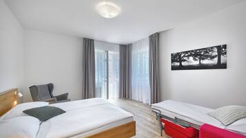 Quarto de hotel moderno com cama de madeira, sofá vermelho, poltrona cinza e uma janela grande com cortinas leves.