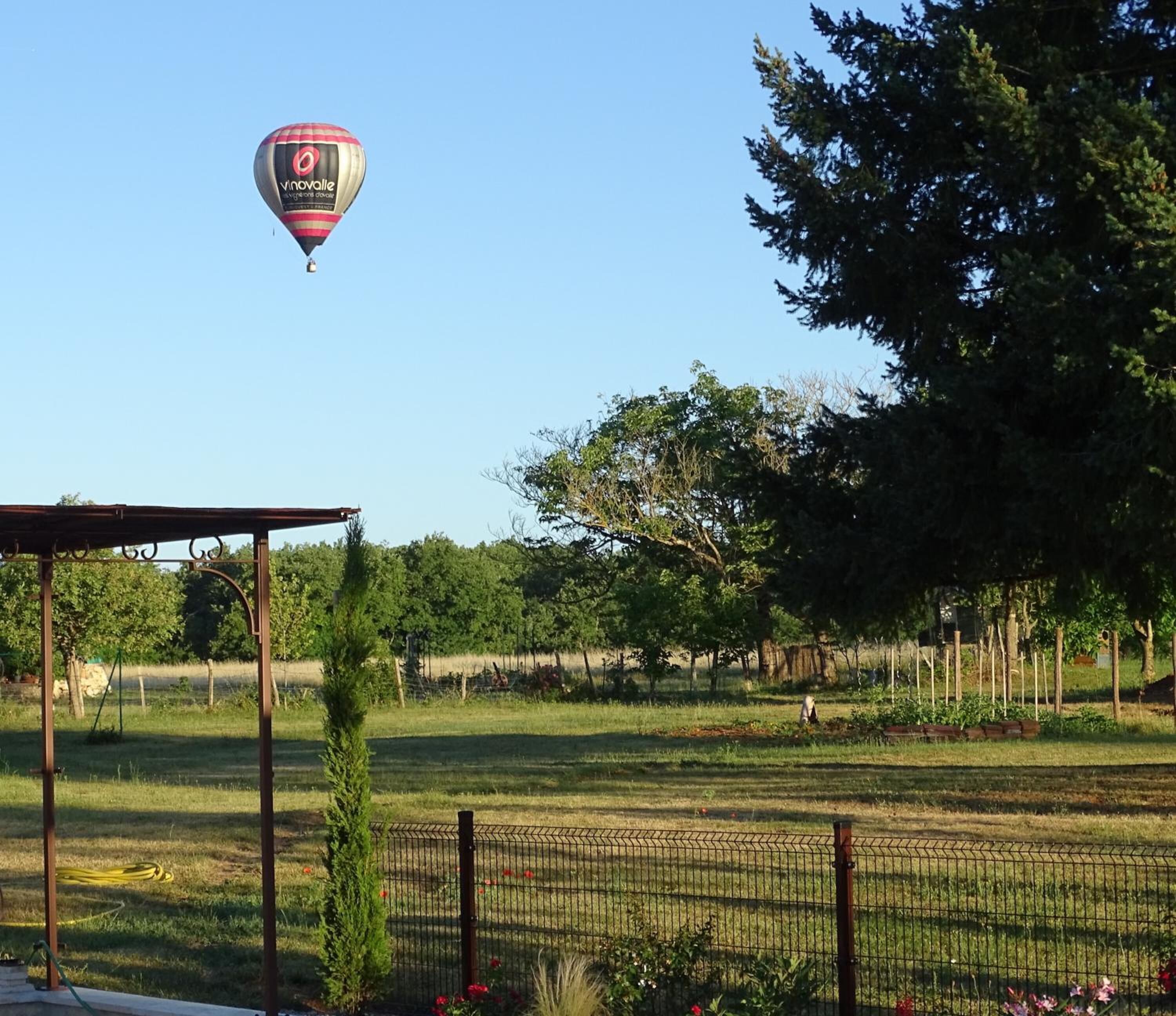 Um balão de ar quente no céu sobre um campo cercado com árvores e uma pérgola.