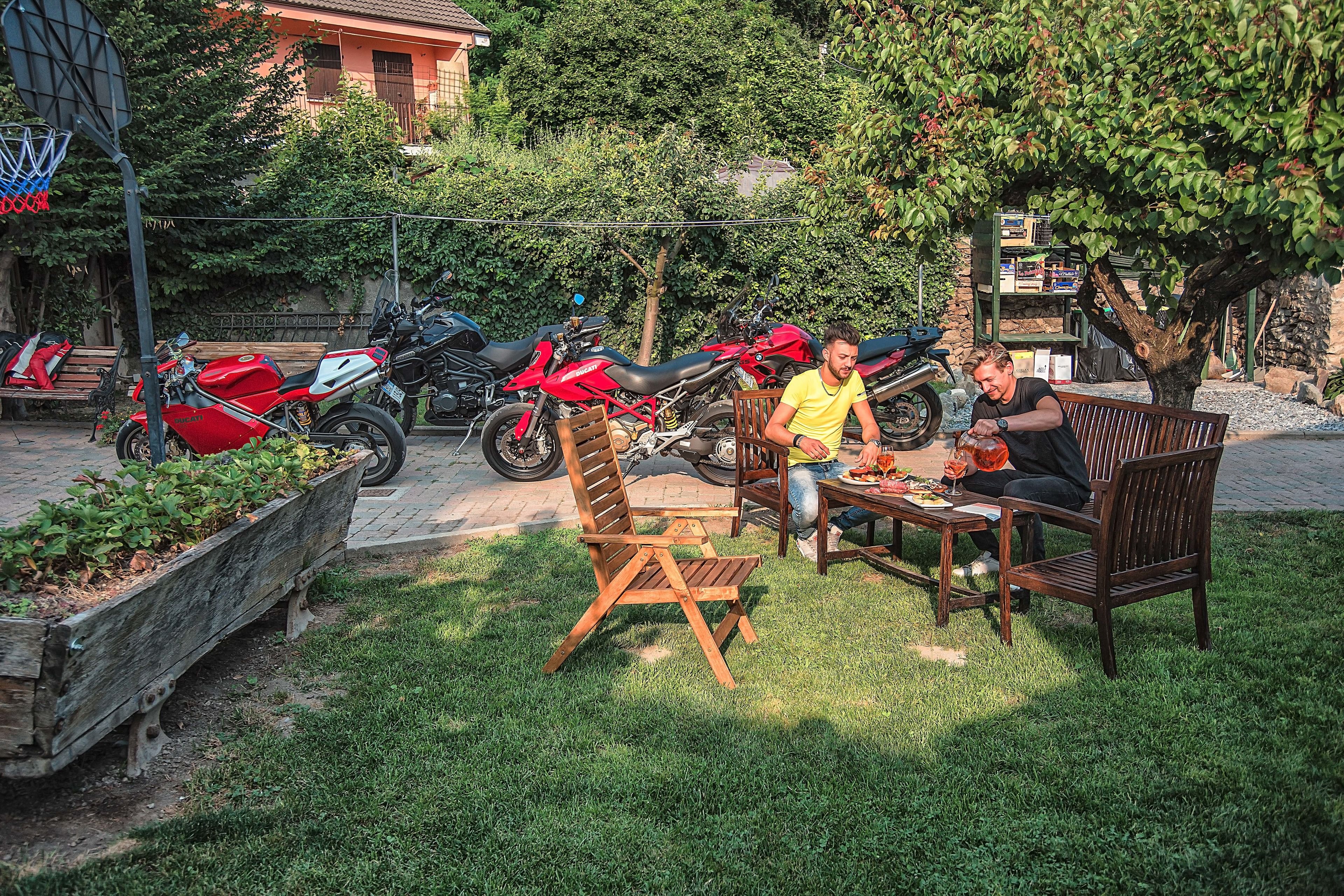Um pátio com um banco e mesa de madeira, cercado por vegetação e motocicletas estacionadas.