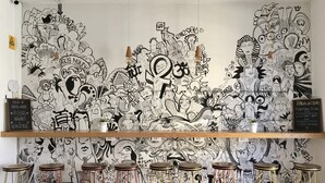 Restaurante com um mural na parede com várias ilustrações e texto. É possível ver banquetas e um menu em lousa.
