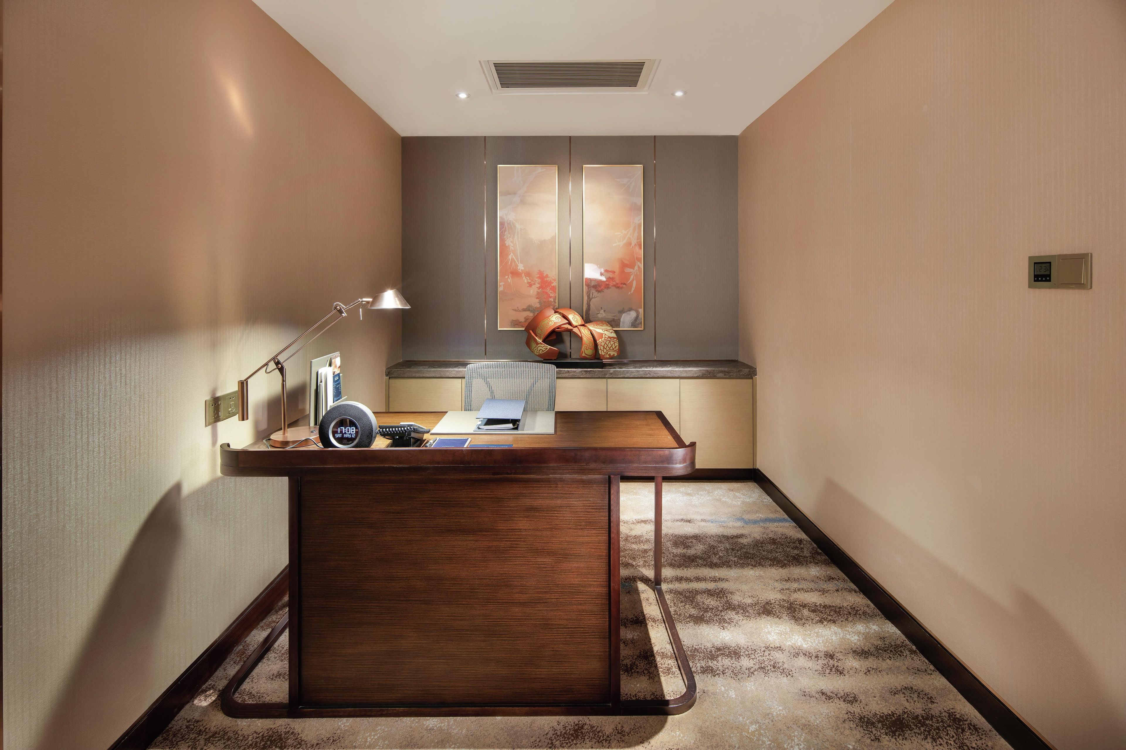 Quarto de hotel com escrivaninha de madeira, luminária, relógio e uma grande pintura na parede.