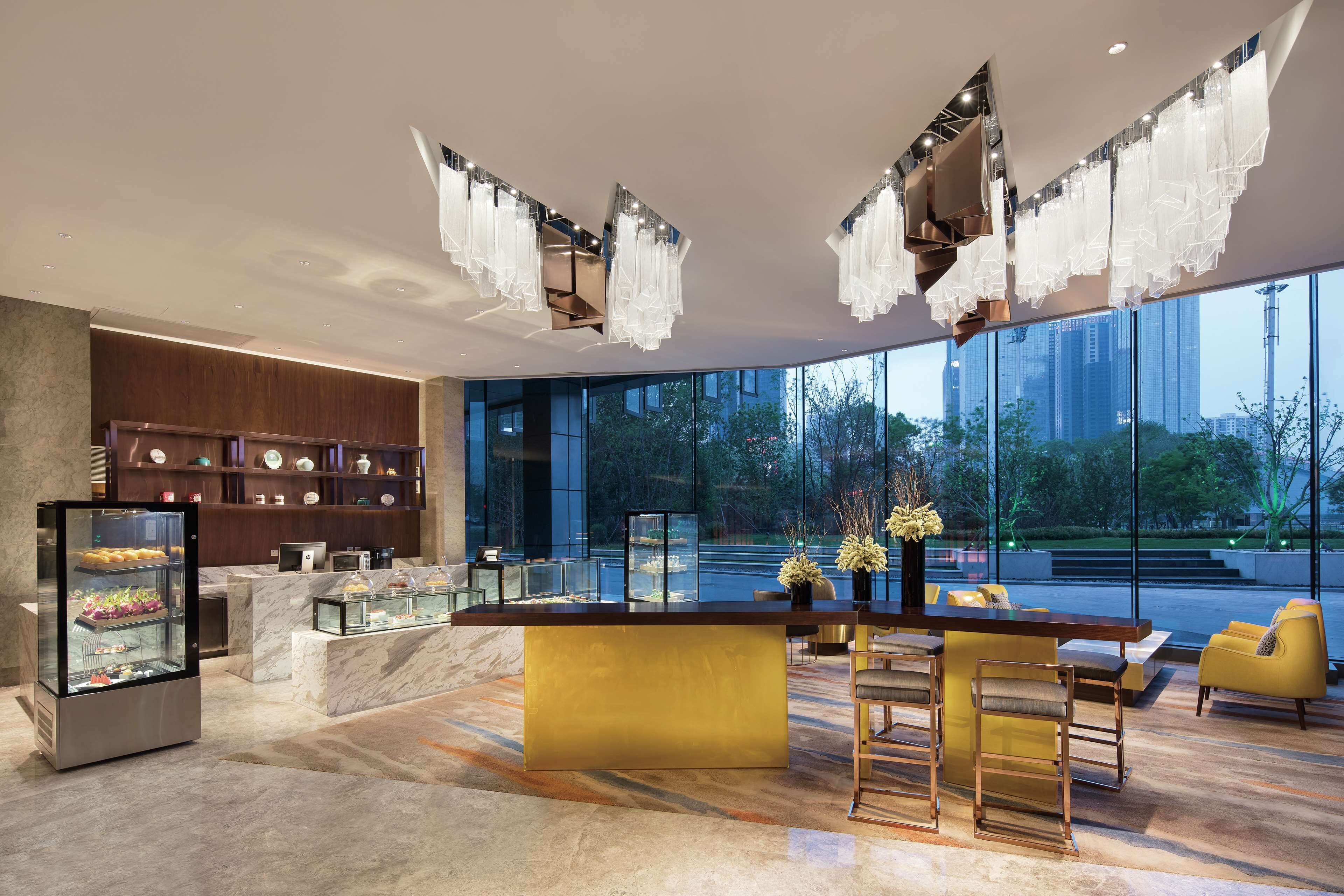 Um saguão de hotel moderno com uma recepção de mármore, uma vitrine de vidro com doces e uma grande janela com vista para a cidade.