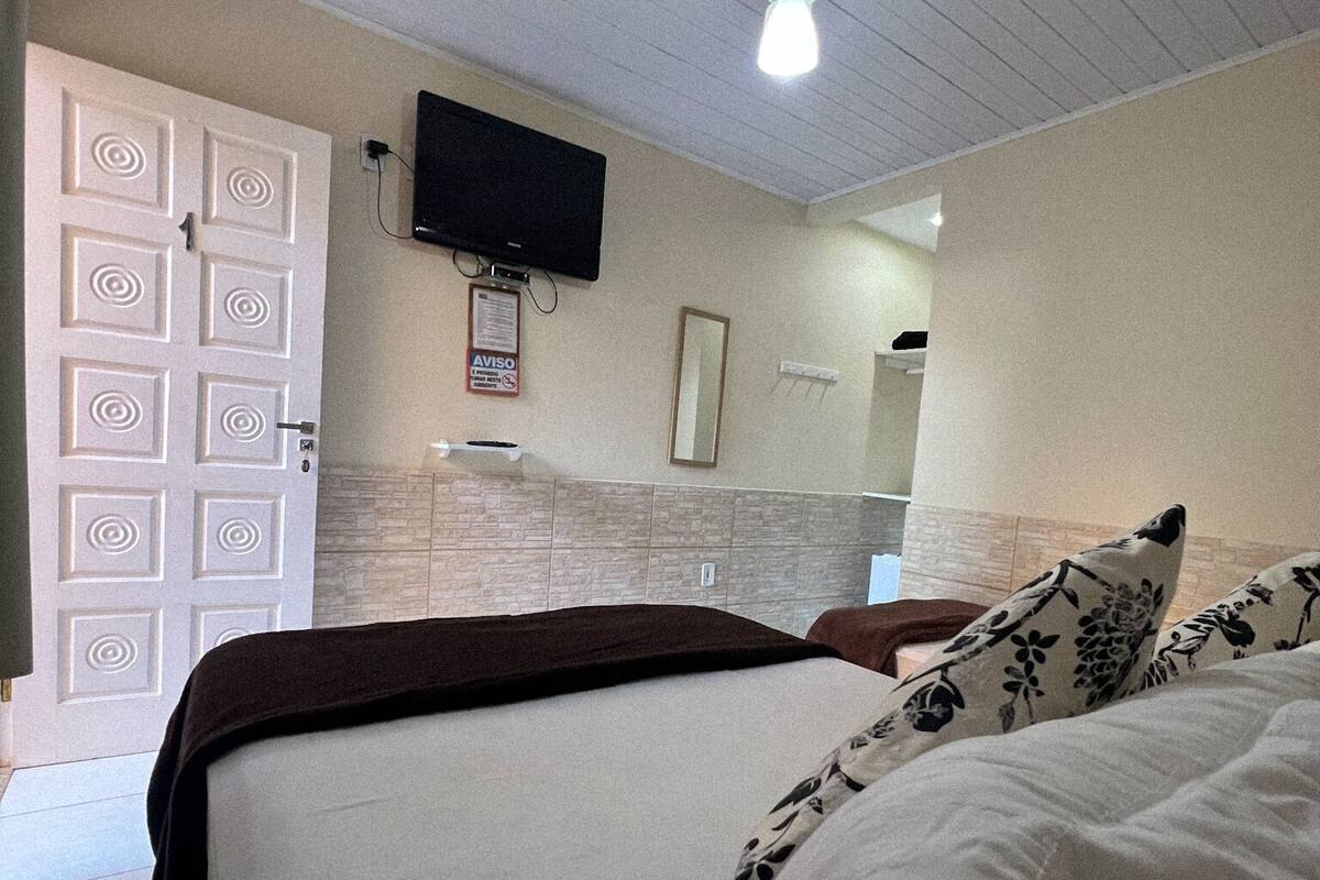 Quarto de hotel com cama, ventilador de teto, televisão de tela plana, porta com olho de fechadura e espelho.