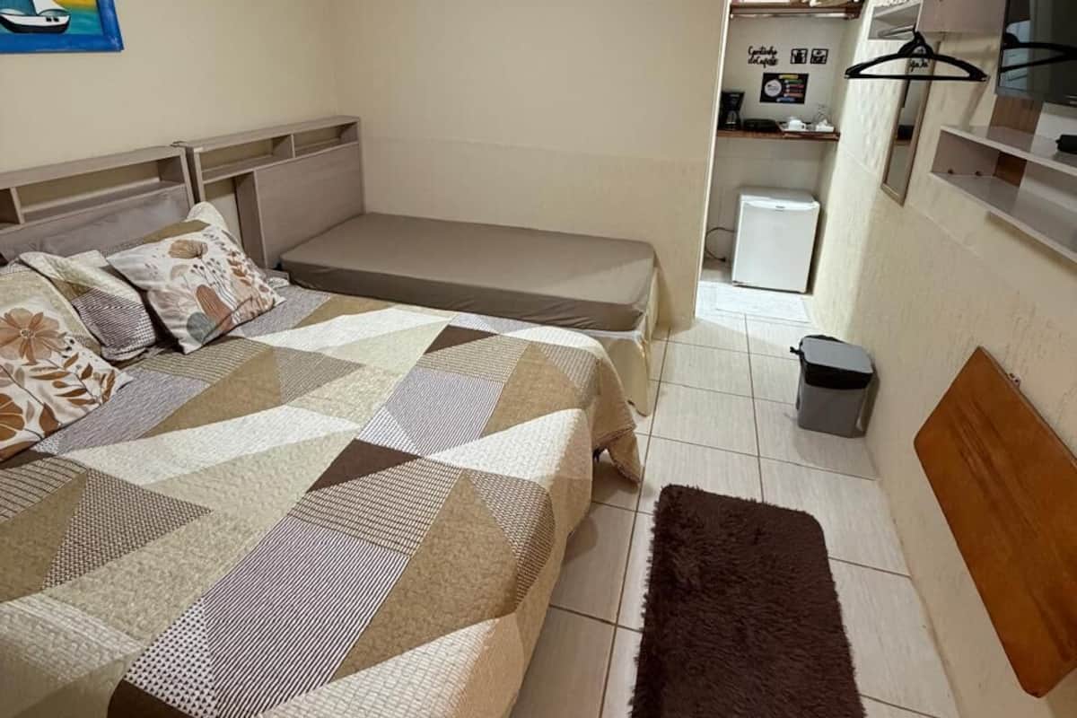 Quarto compacto com cama, pequeno espaço de cozinha, ventoinha no teto e lixeira.
