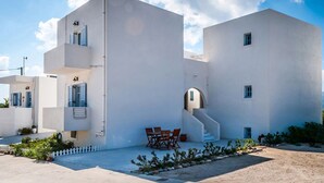 Um edifício moderno e branco com telhado plano, uma pequena área externa com mesas e cadeiras, e uma escadaria que leva a um andar superior.