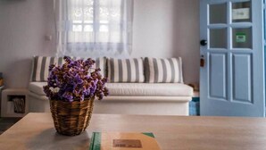 Um quarto com uma cesta de vime cheia de flores roxas sobre uma mesa, um sofá branco com almofadas listradas e uma porta azul com janela.