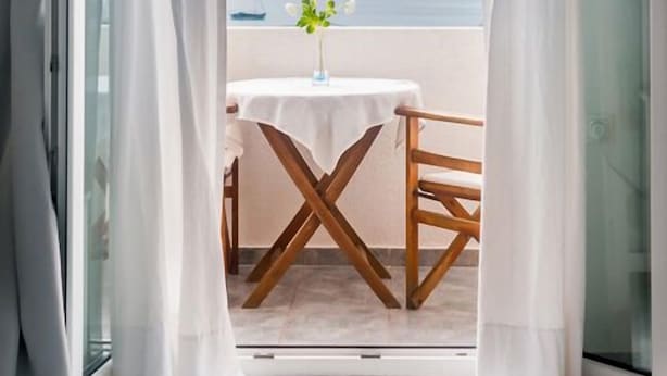 Uma varanda com mesa e cadeiras, um vaso com flores e vista para o mar e as montanhas.