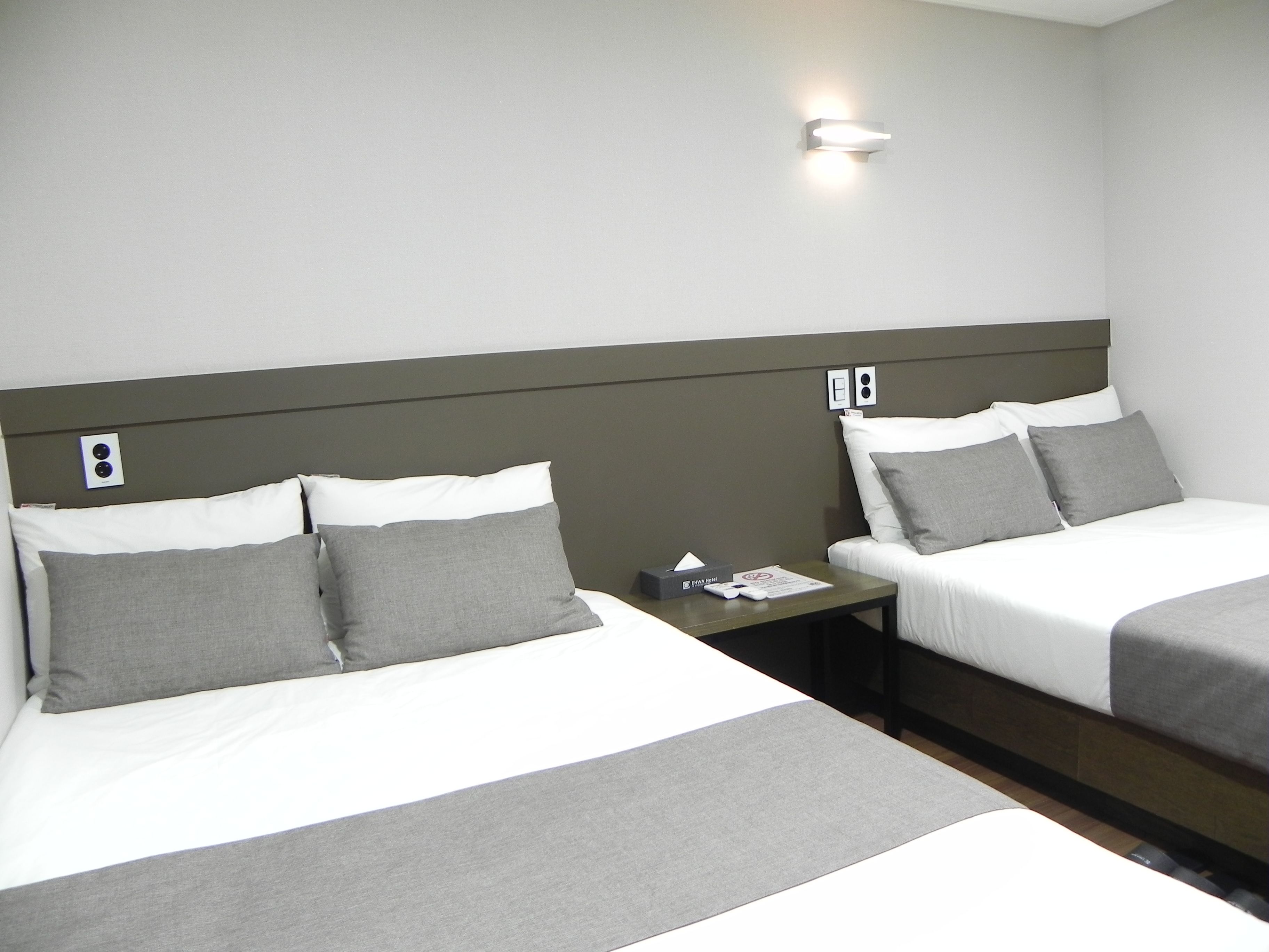 Quarto de hotel com duas camas, cada uma com roupa de cama e almofadas cinzas, uma mesinha de cabeceira com uma caixa de lenços, e um candeeiro fixado na parede.