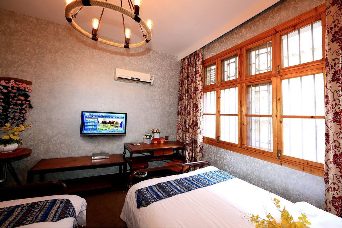 Quarto com duas camas, uma escrivaninha, televisão e uma janela grande com cortinas.