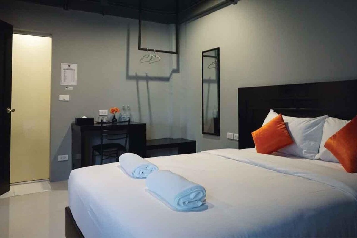 Quarto de hotel com uma cama, duas toalhas enroladas, uma escrivaninha com cadeira e um espelho na parede.