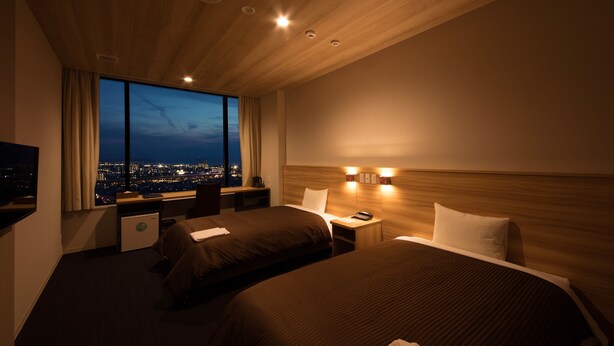 Quarto de hotel com duas camas, uma escrivaninha, uma cadeira, um criado-mudo e vista para a cidade à noite.