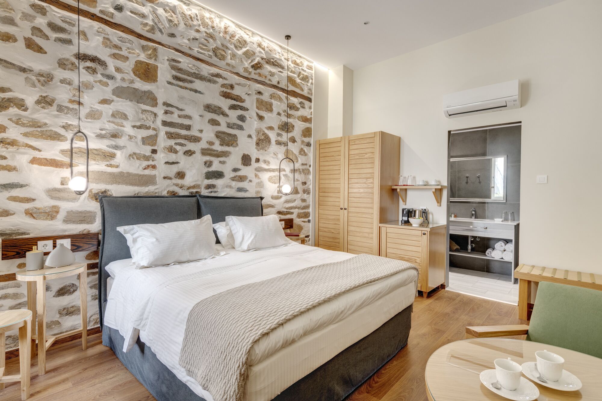 Quarto moderno com parede de pedra, cama grande com roupa de cama cinzenta, armário de madeira, pequena mesa com xícaras e banheiro com espelho e pia.