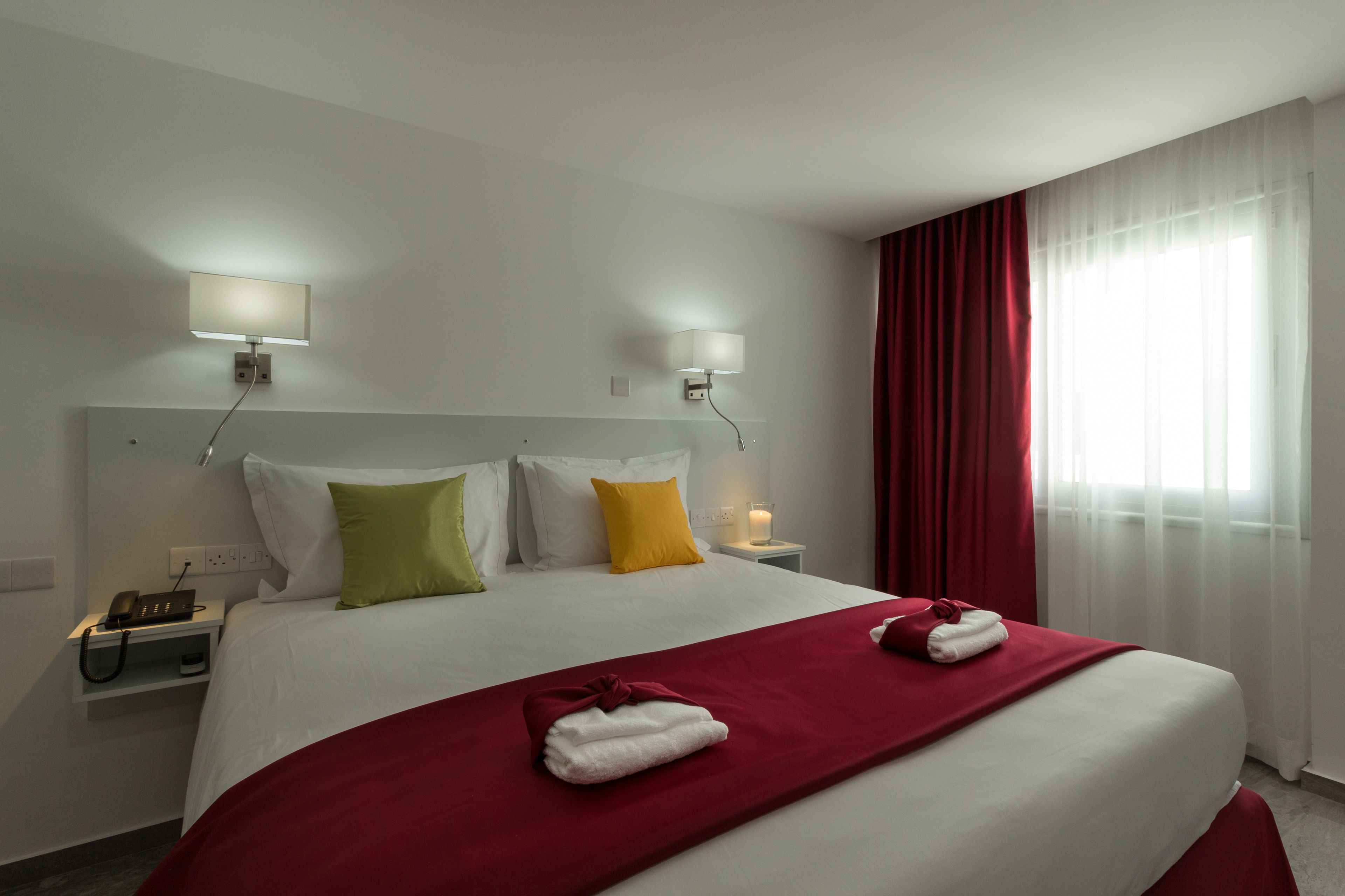 Quarto de hotel com uma cama grande, duas luminárias de cabeceira, uma mesa de cabeceira com telefone e uma janela com cortinas vermelhas.