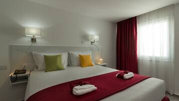 Quarto de hotel com uma cama grande, duas luminárias de cabeceira, uma mesa de cabeceira com telefone e uma janela com cortinas vermelhas.