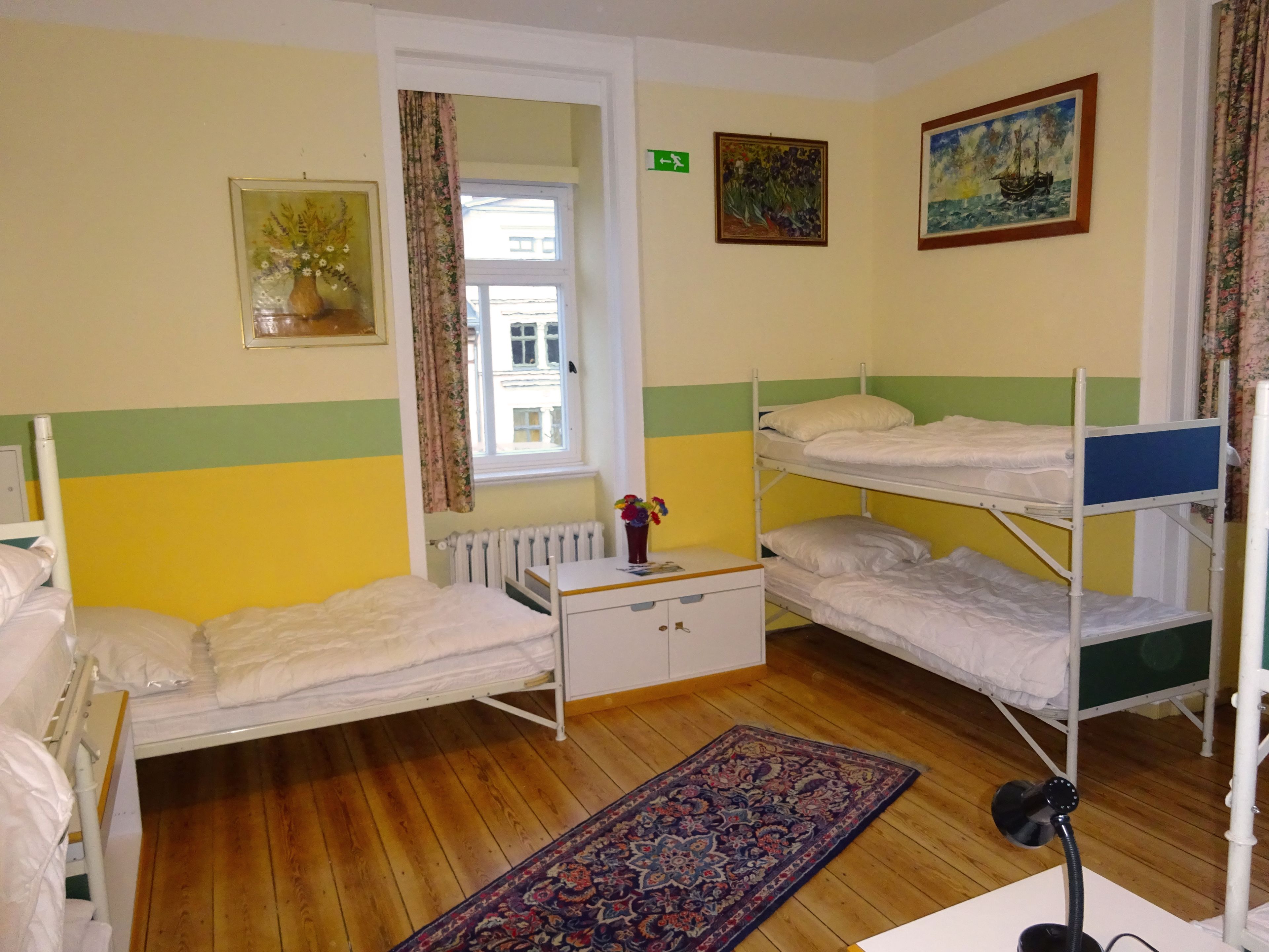 Um dormitório com beliches, um radiador, uma janela com cortinas, uma escrivaninha com luminária e um tapete.