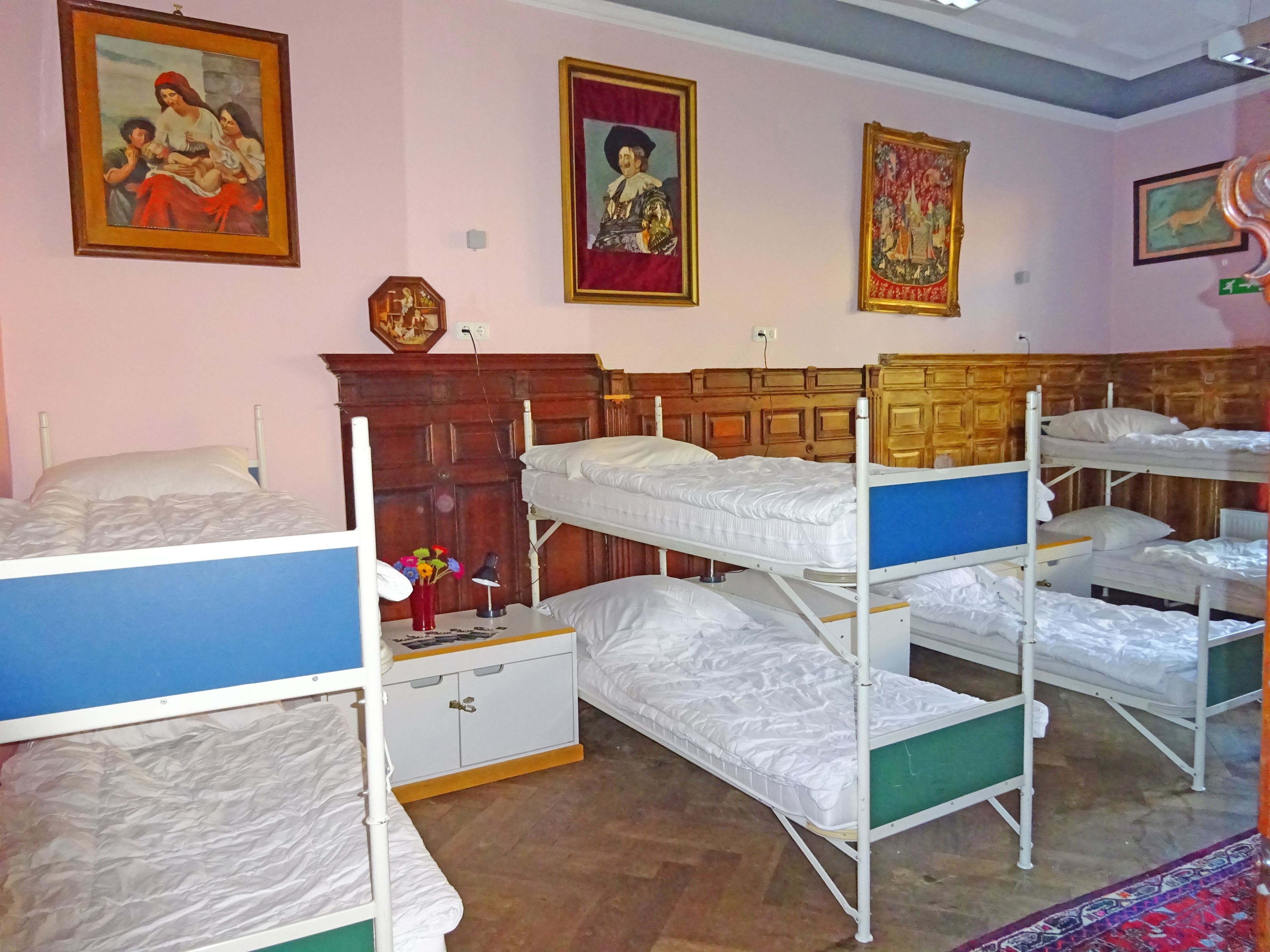 Um dormitório com beliches, cabeceira de madeira, escrivaninha com luminária e pinturas religiosas nas paredes.