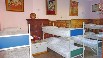 Um dormitório com beliches, cabeceira de madeira, escrivaninha com luminária e pinturas religiosas nas paredes.